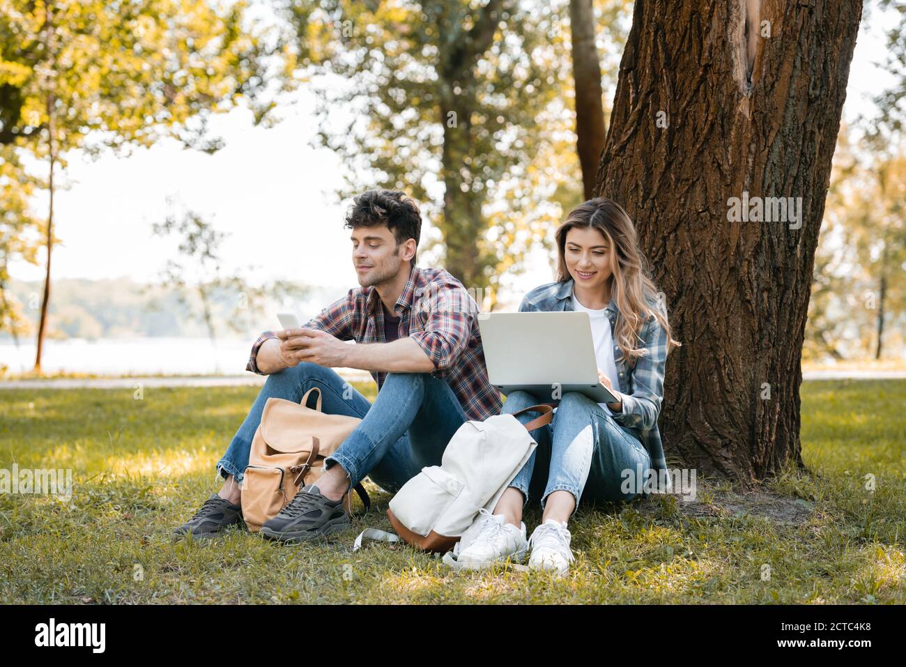 couple using laptop and smartphone while sitting under tree trunk Stock ...