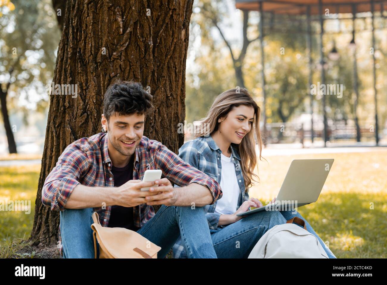 joyful couple using gadgets while sitting under tree trunk Stock Photo ...