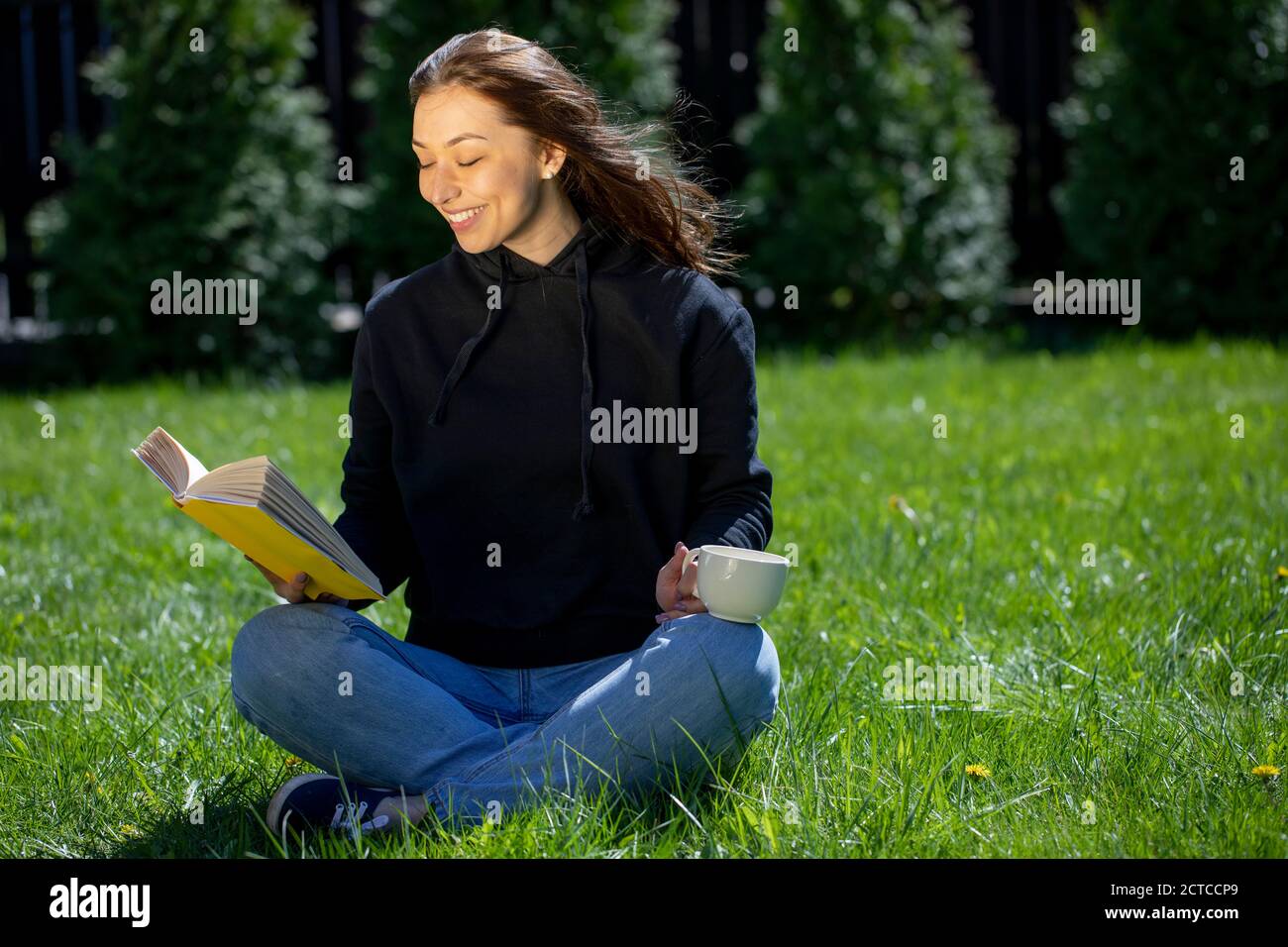 Woman is sitting on the lawn in the garden reading a book and drinking ...