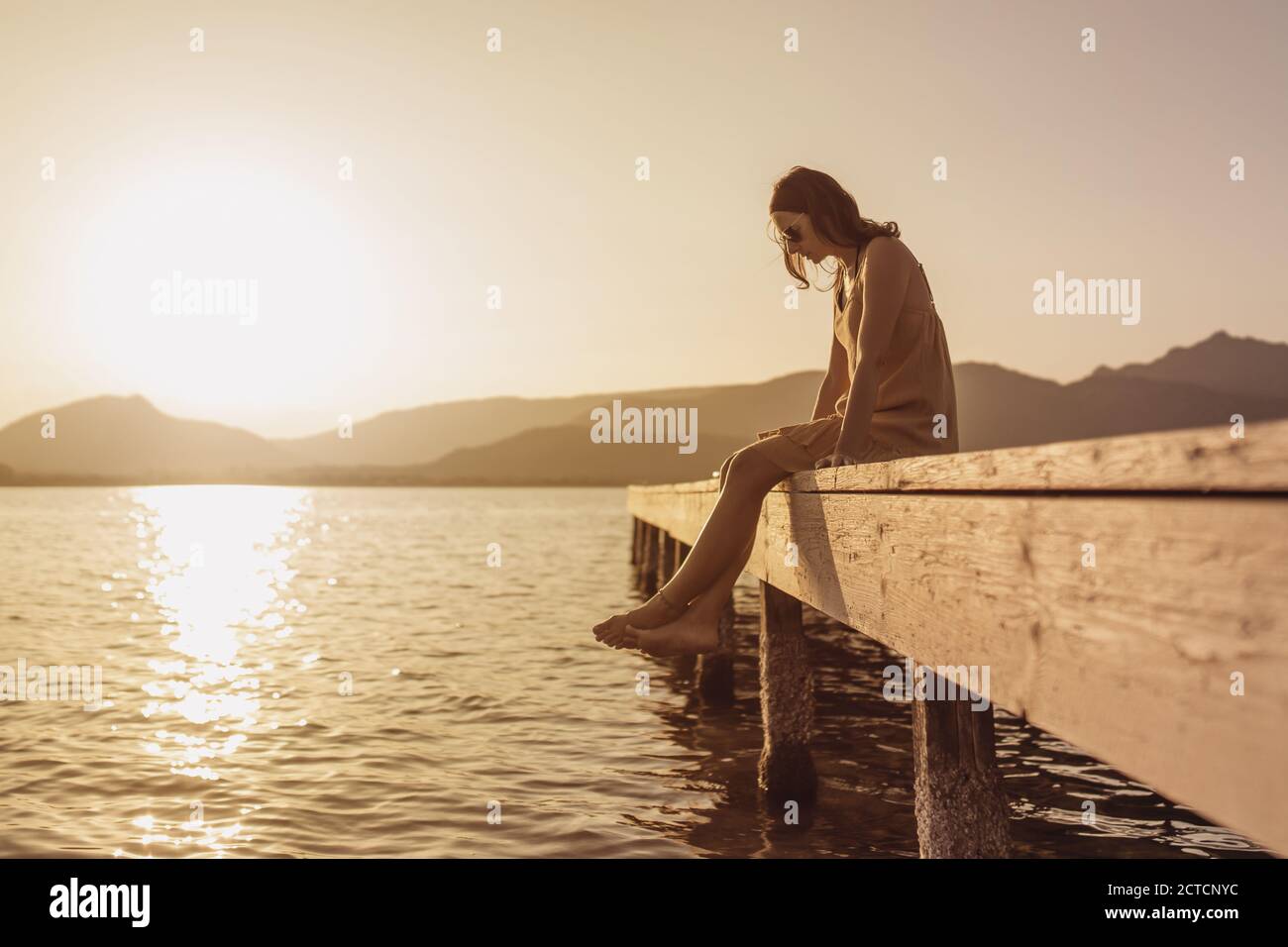 Pensive single Caucasian pretty young woman sitting on a pier of a lake ...