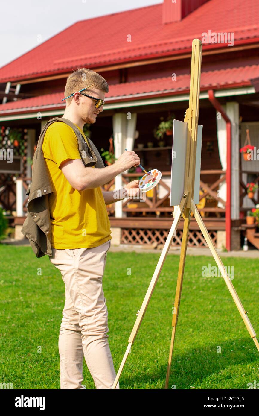 young man paints painting outside his country house holding palette of ...
