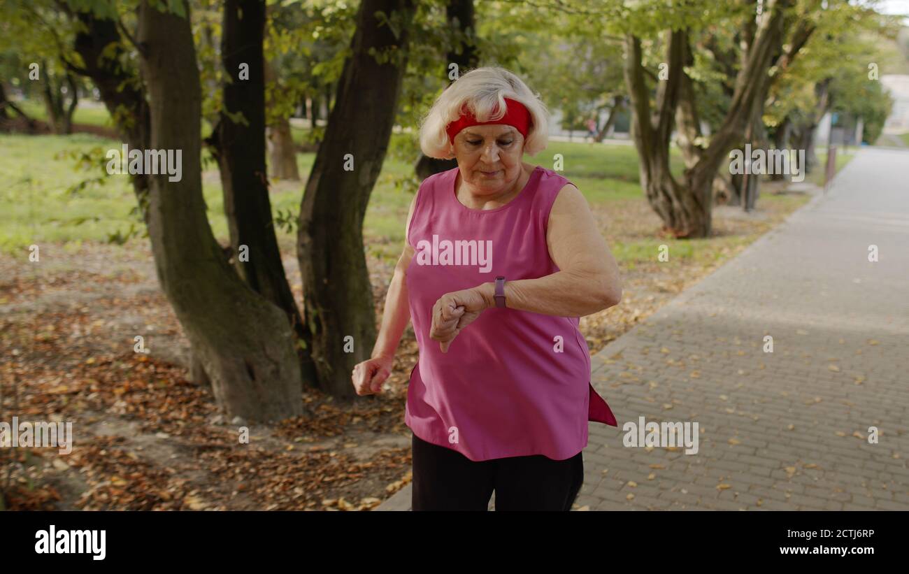 Female senior person running along the road in city park. Mature runner ...