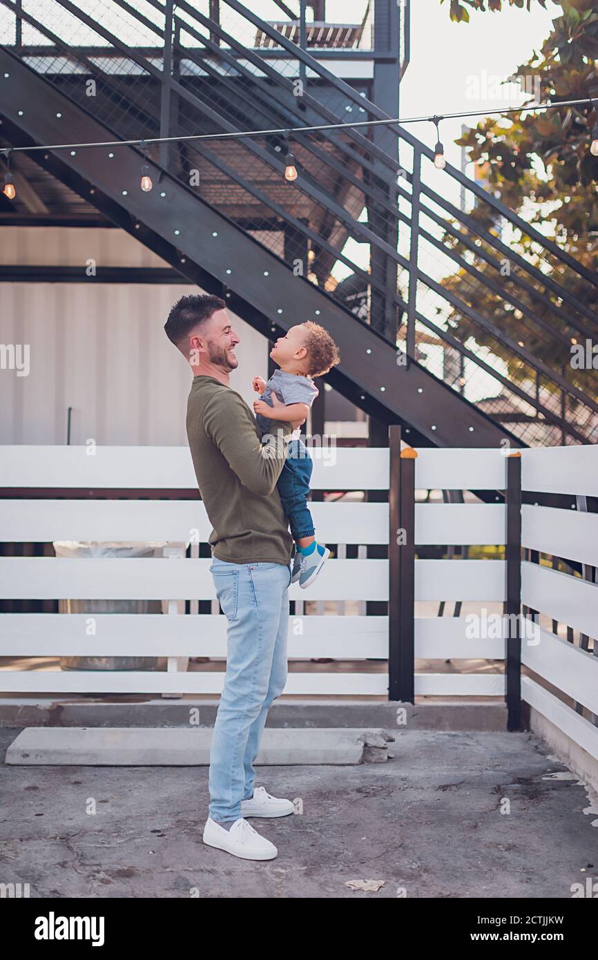 Dad laughing while holding boy in front of white fence and black steps ...