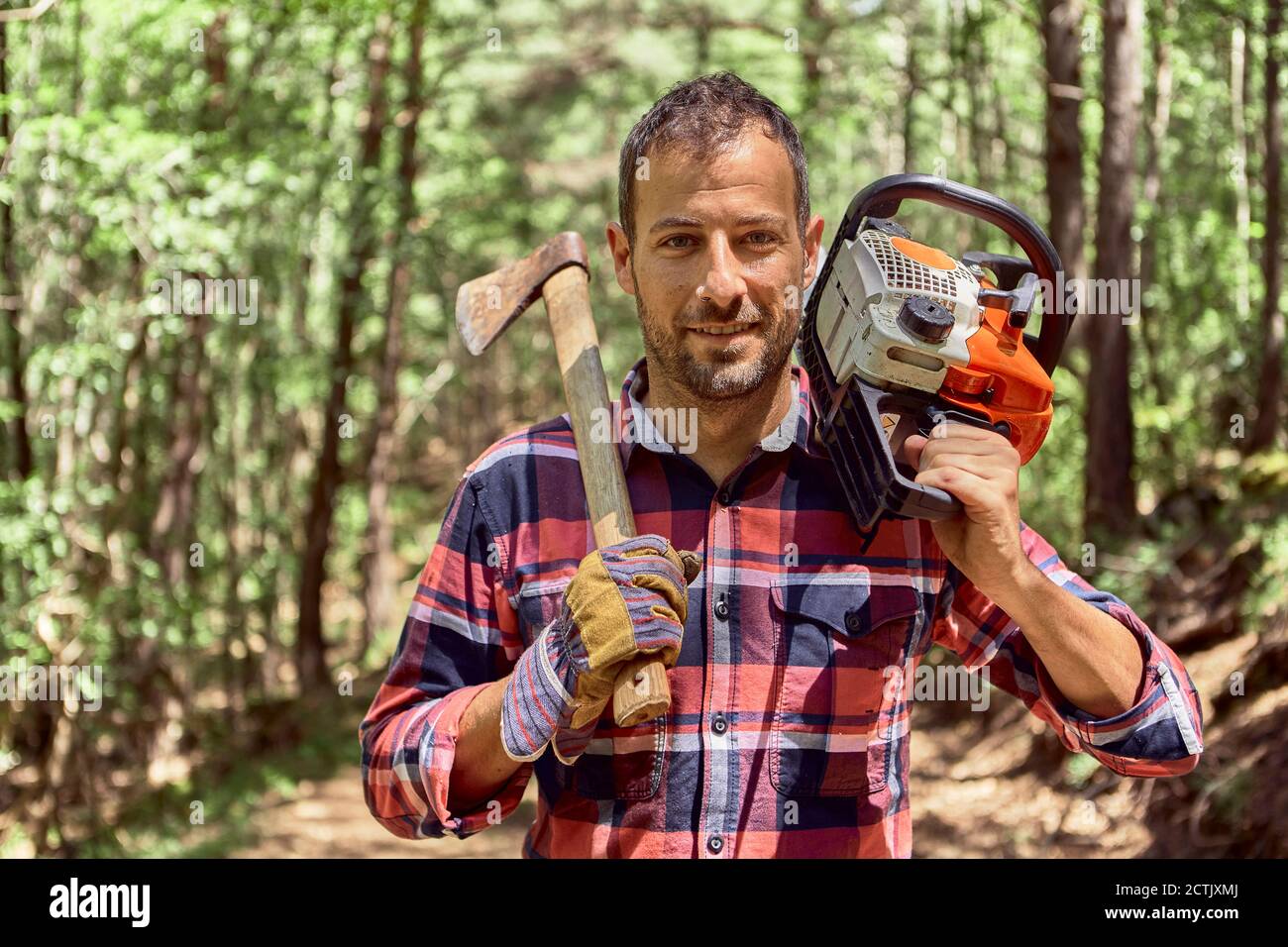 Smiling lumberjack standing with axe and chainsaw in forest Stock Photo ...