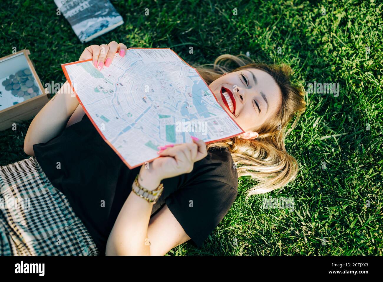 Smiling young woman holding map while lying on grassy land in park ...