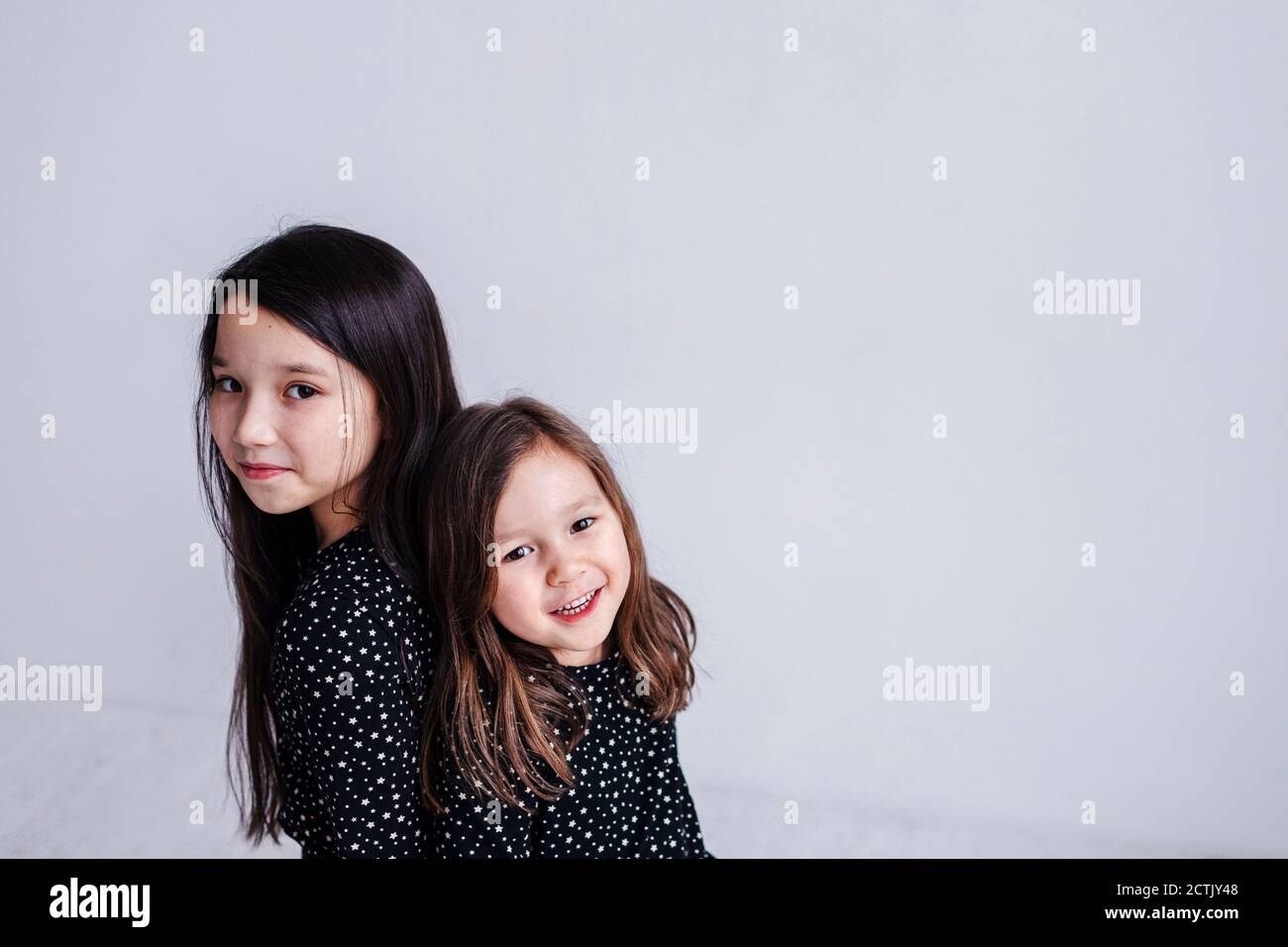 Cute sisters standing back to back against white background in studio ...