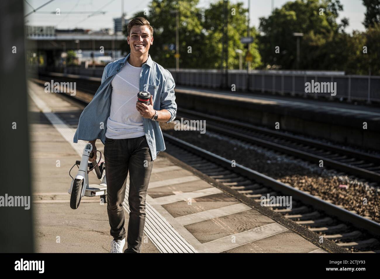 Smiling young man carrying push scooter while walking on railroad ...