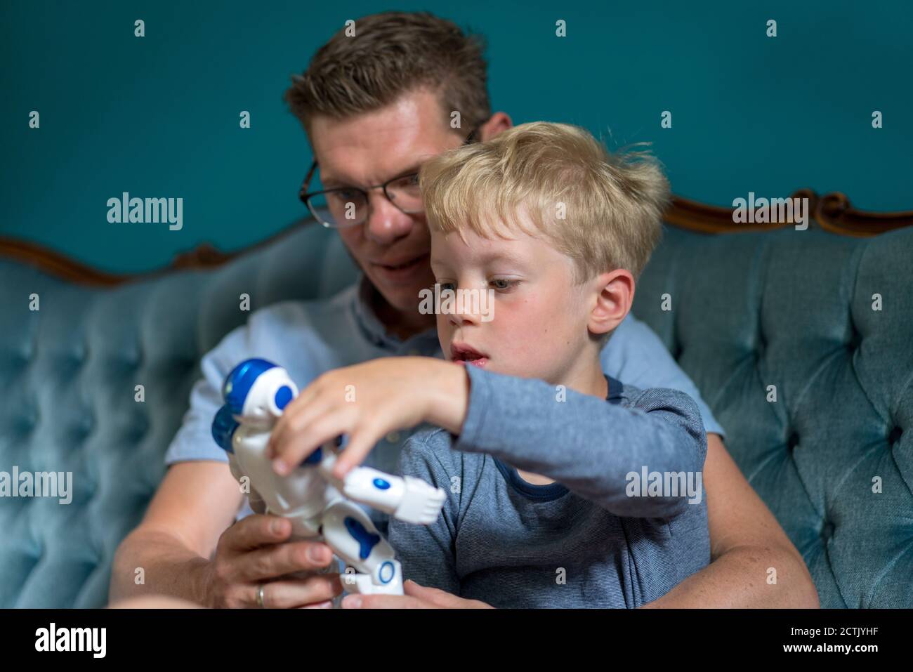 Blond boy looking at robot held by father while sitting on sofa in ...