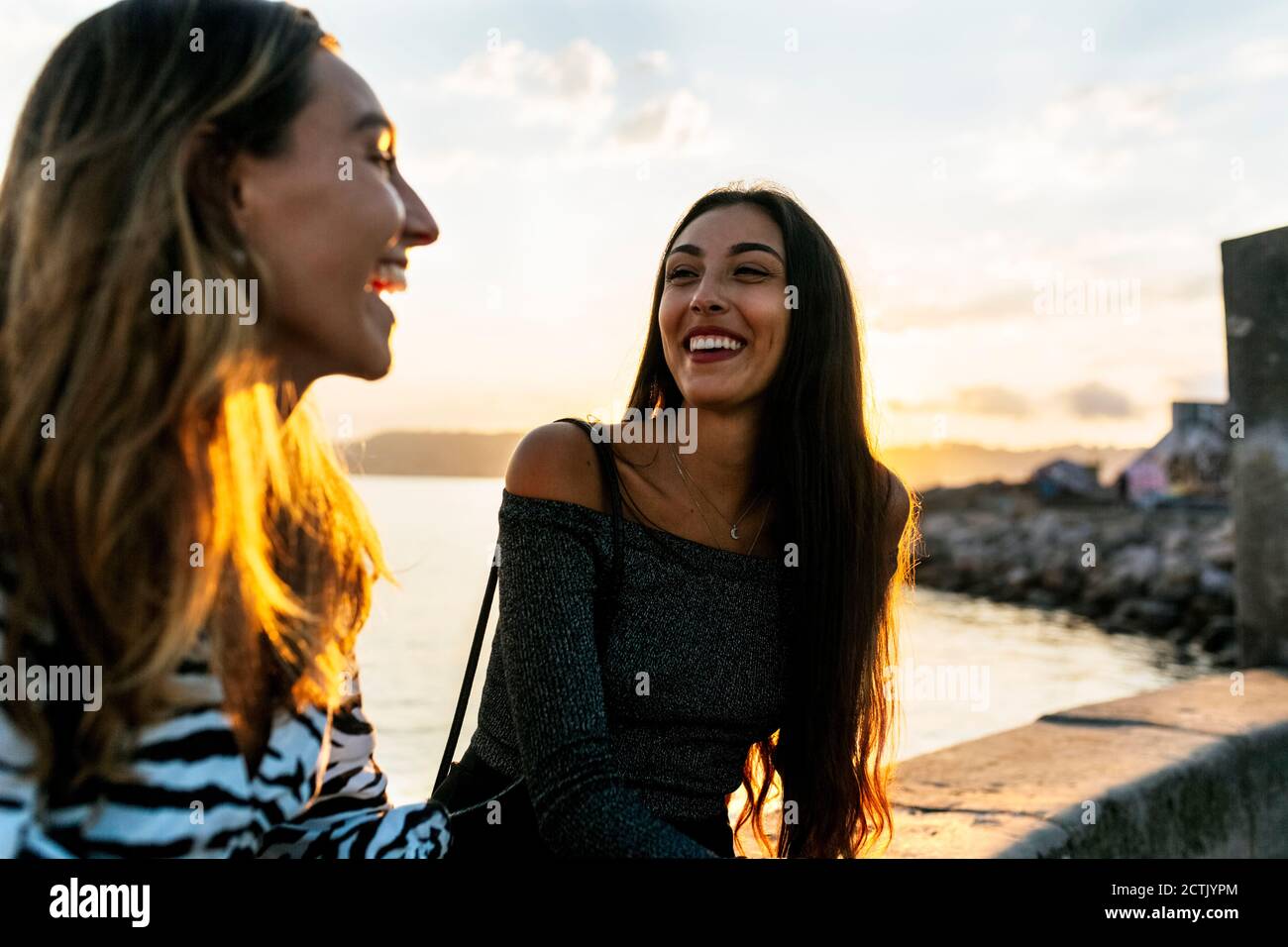 Happy friends talking while sitting on retaining wall promenade during ...