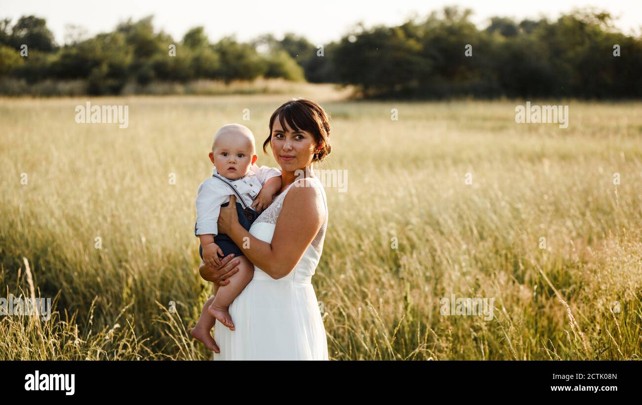 Bride carrying baby boy while standing at field during sunset Stock ...