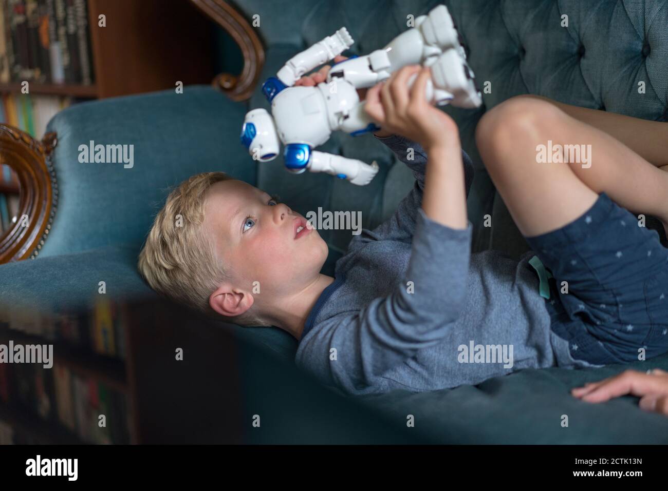 Cute boy playing with robot while lying on sofa in living room at home ...