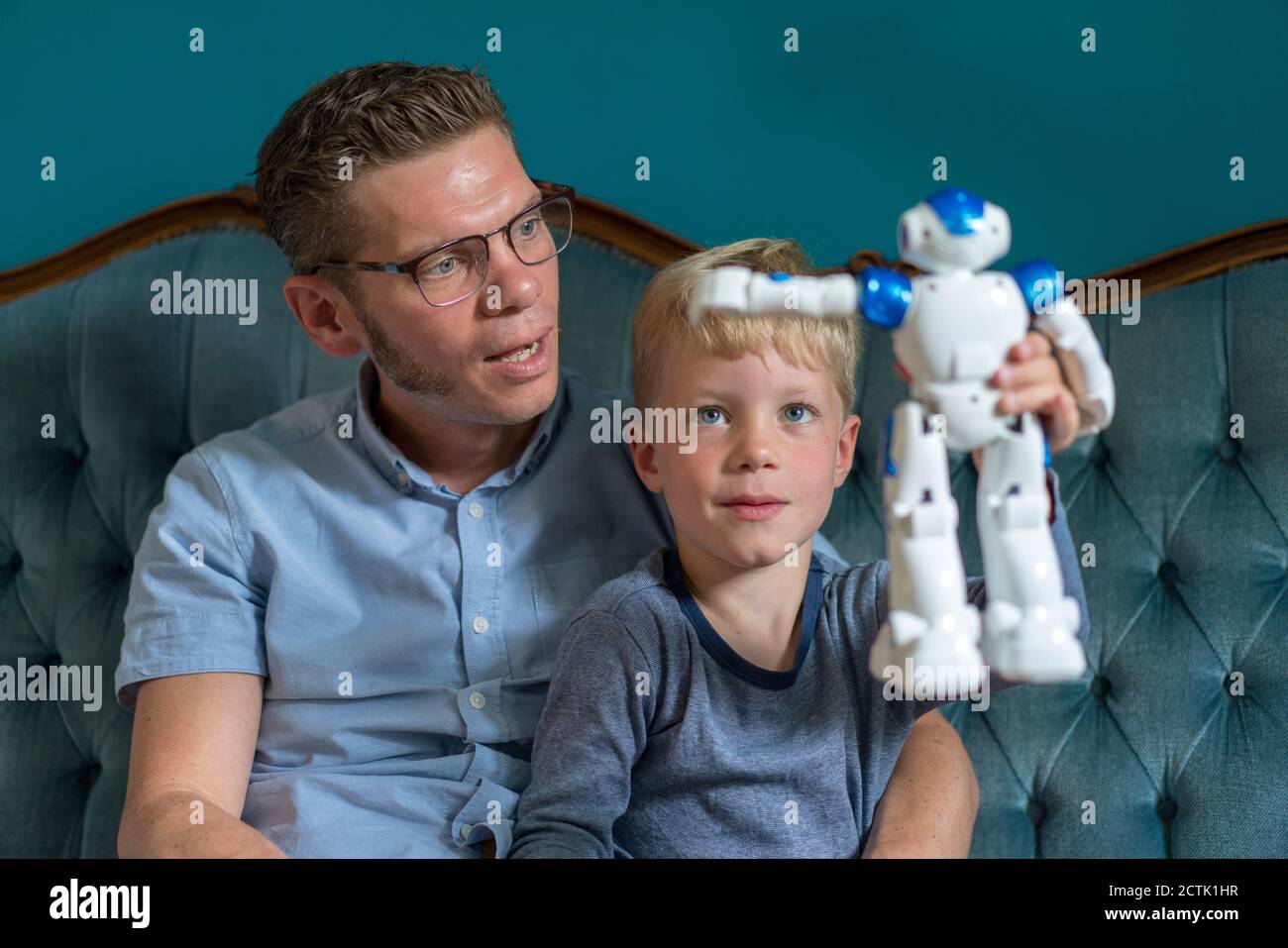 Cute blond boy holding robot while sitting by father on sofa in living ...