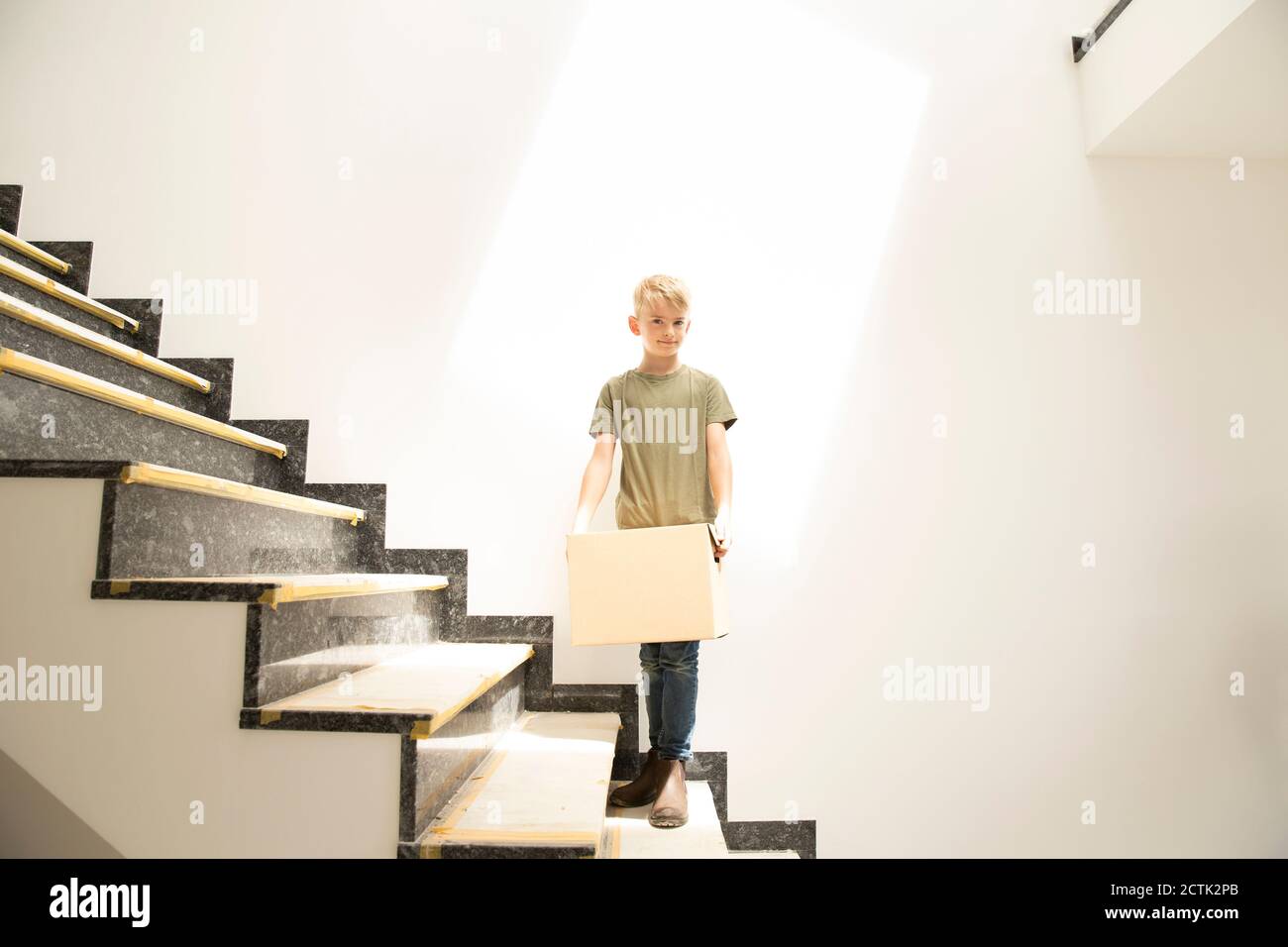 Boy carrying box while standing on staircase against wall in new home ...
