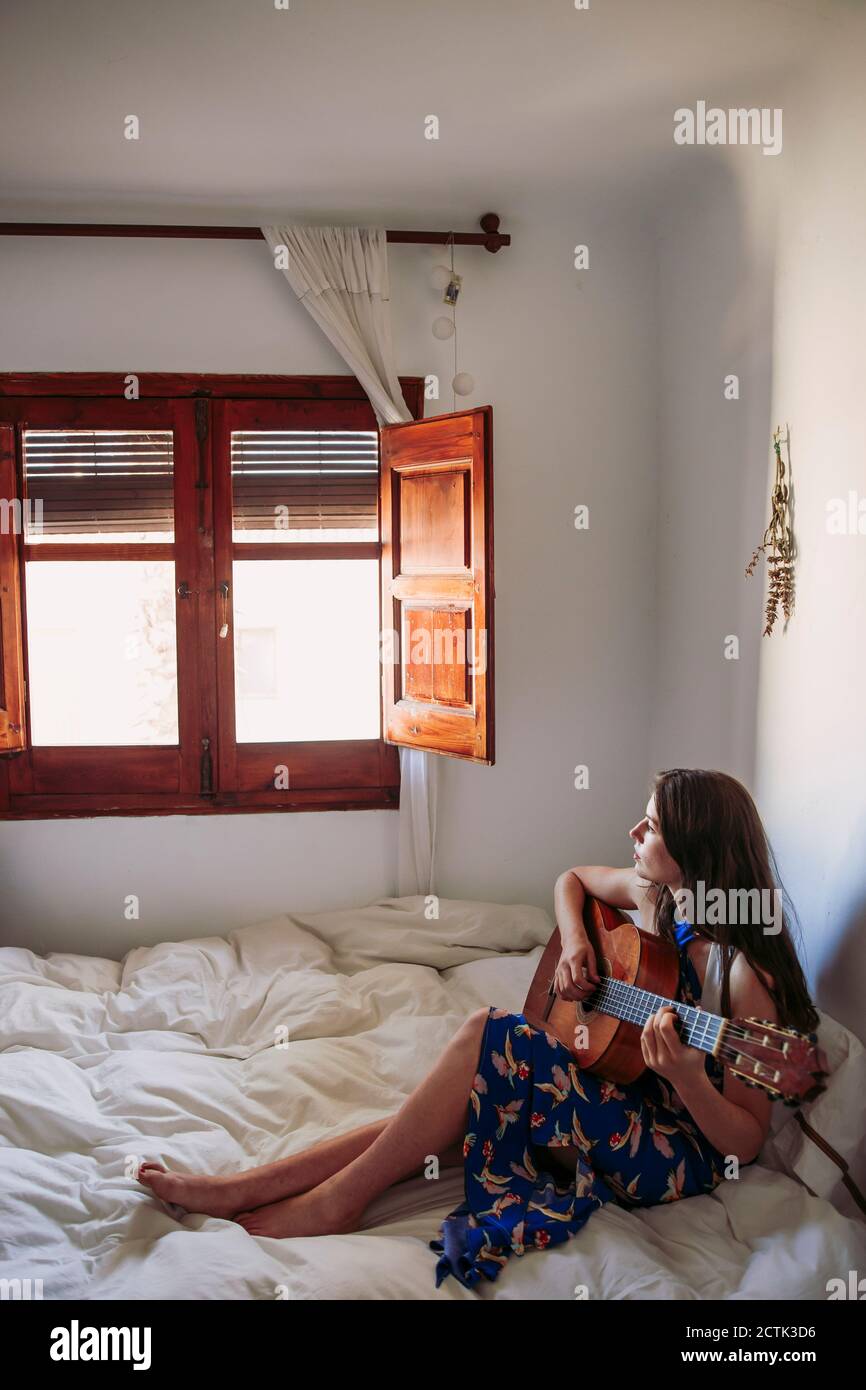 Woman looking through window while practicing guitar in bedroom at home ...
