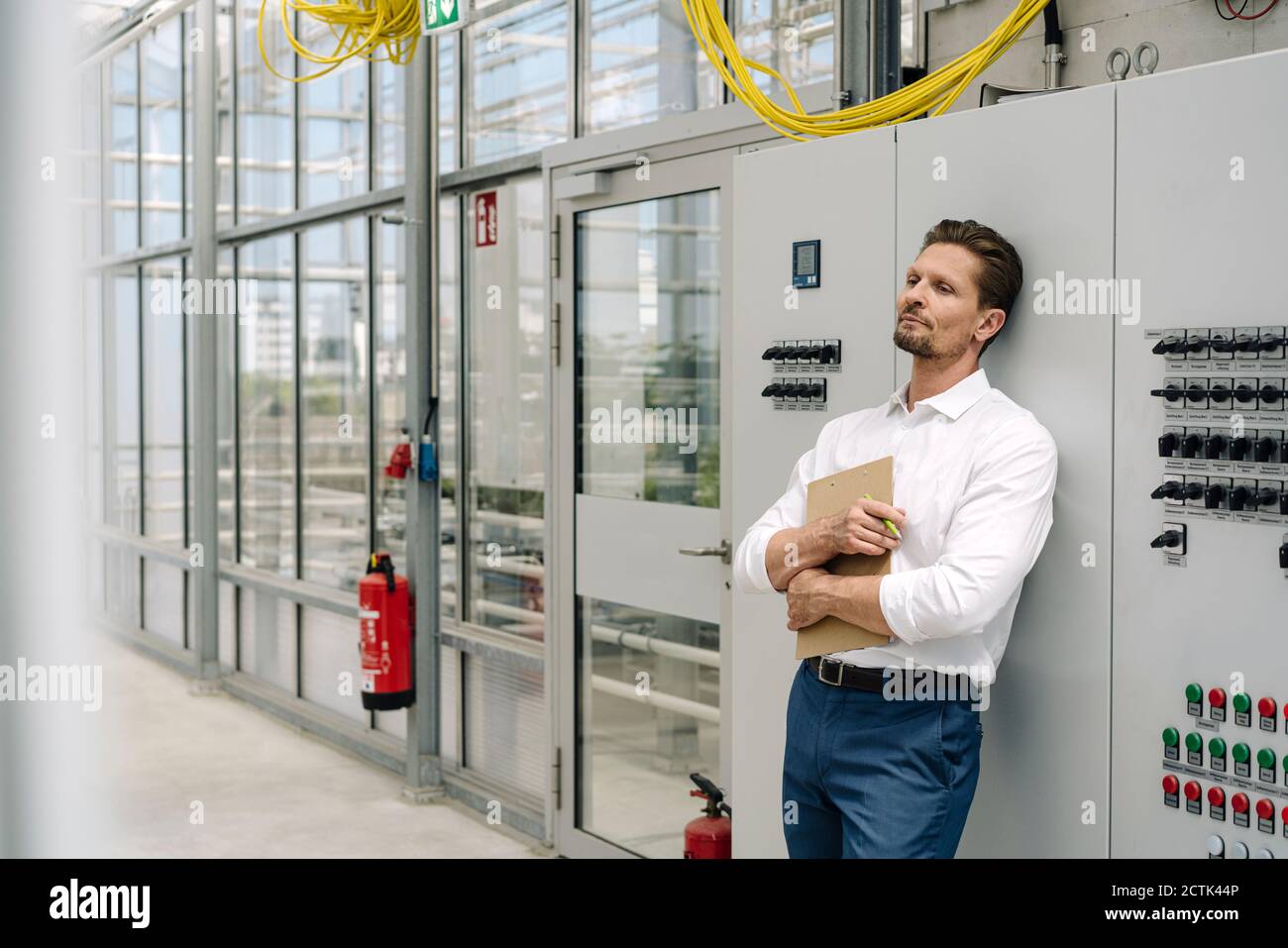 Thoughtful businessman holding clipboard while leaning on control panel ...