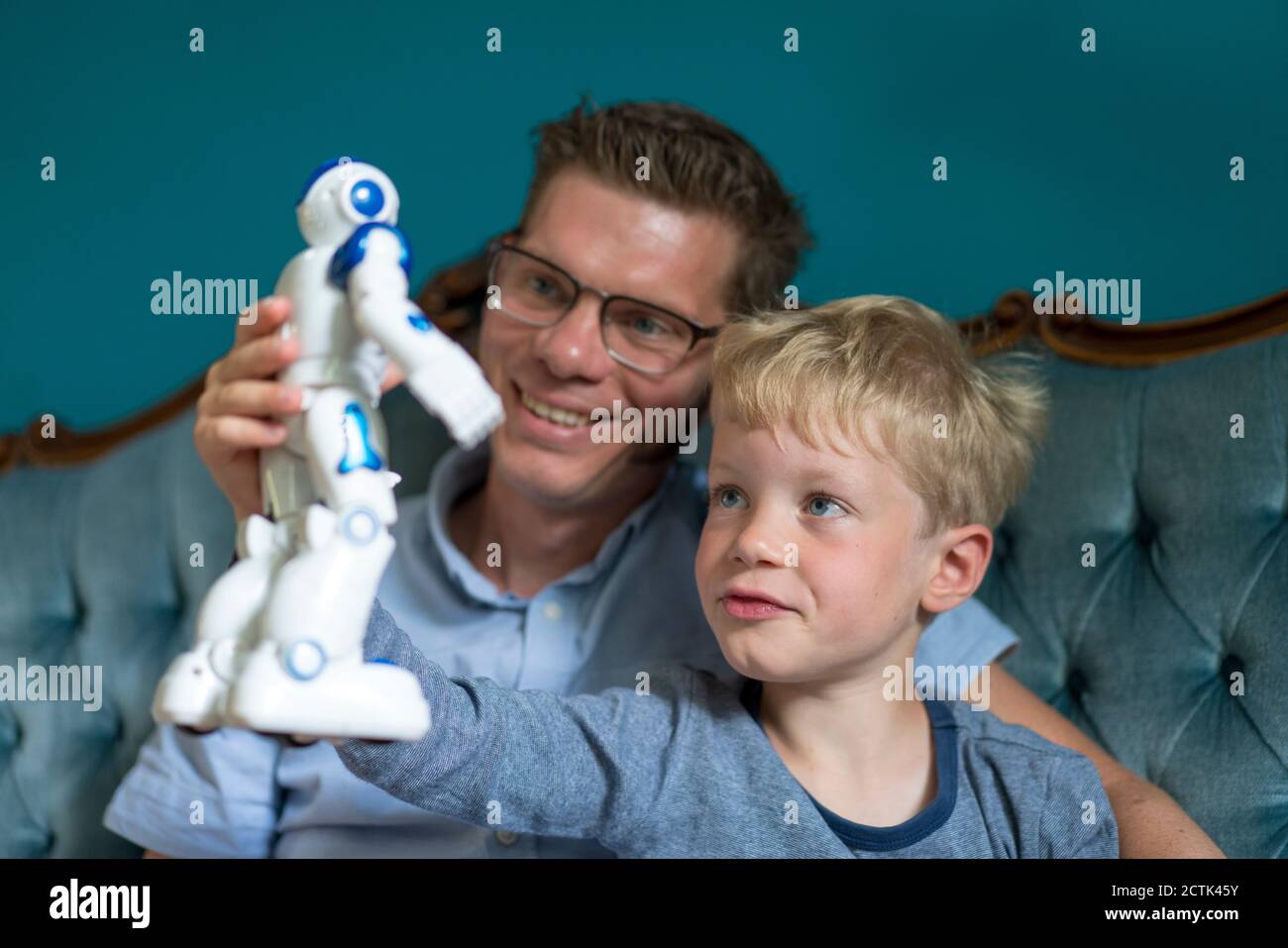 Blond boy looking at robot while sitting by smiling father in living ...