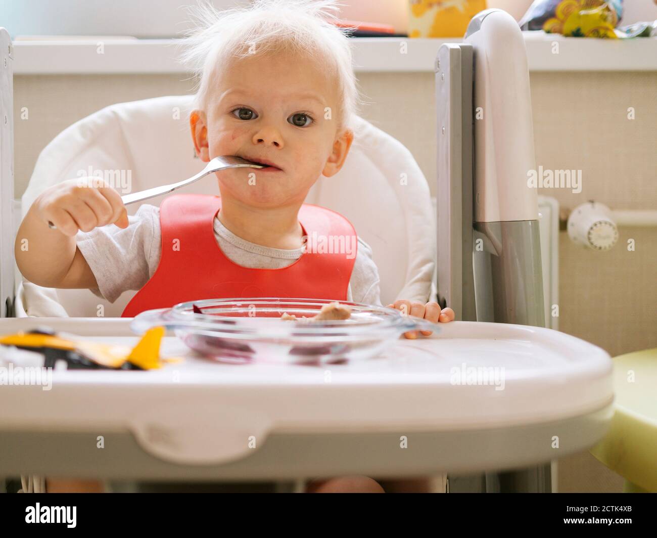Cute little baby biting fork while sitting on high chair in kitchen ...