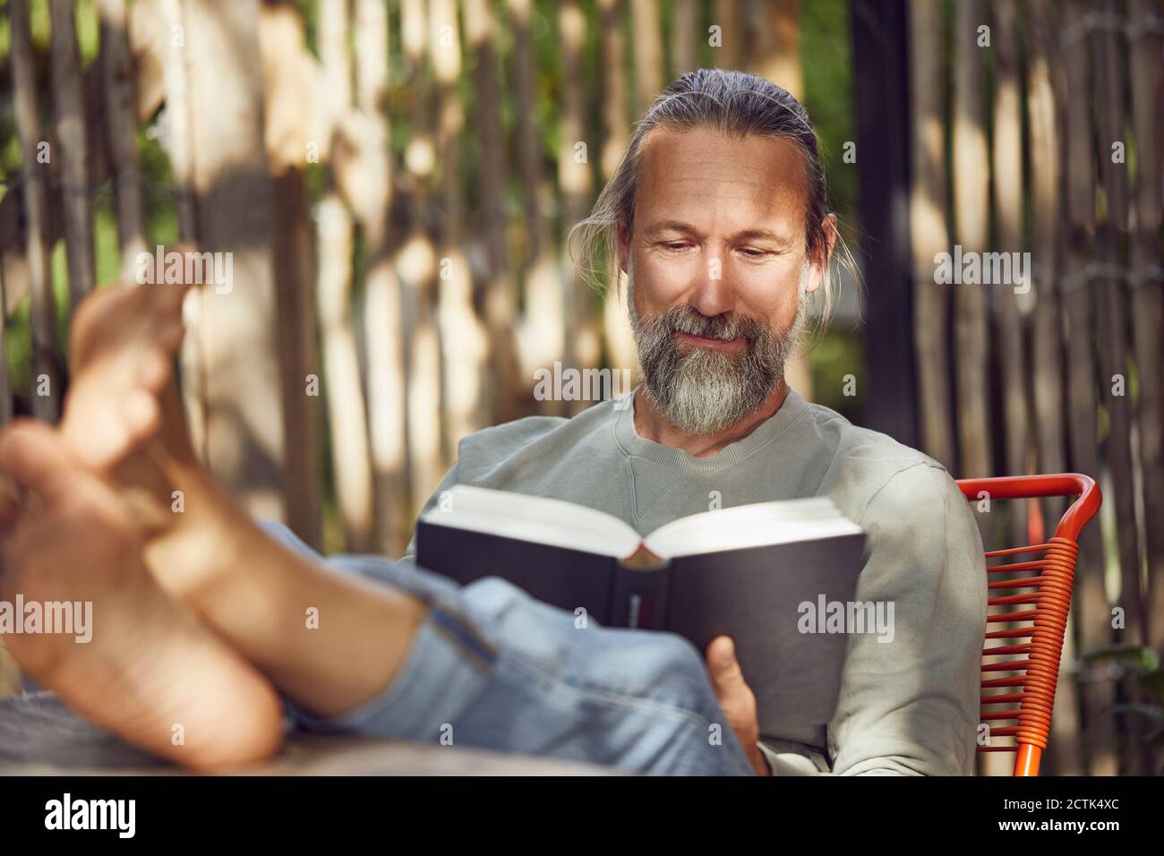Bearded Mature Man Reading Book While Relaxing On Chair In Yard Stock