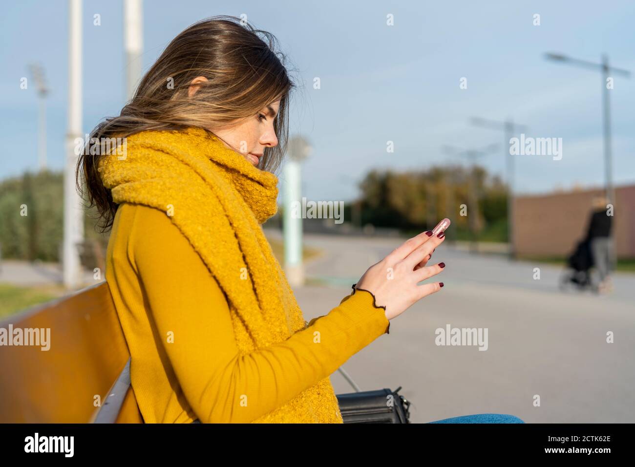 Young woman texting through mobile phone while sitting on bench in park ...