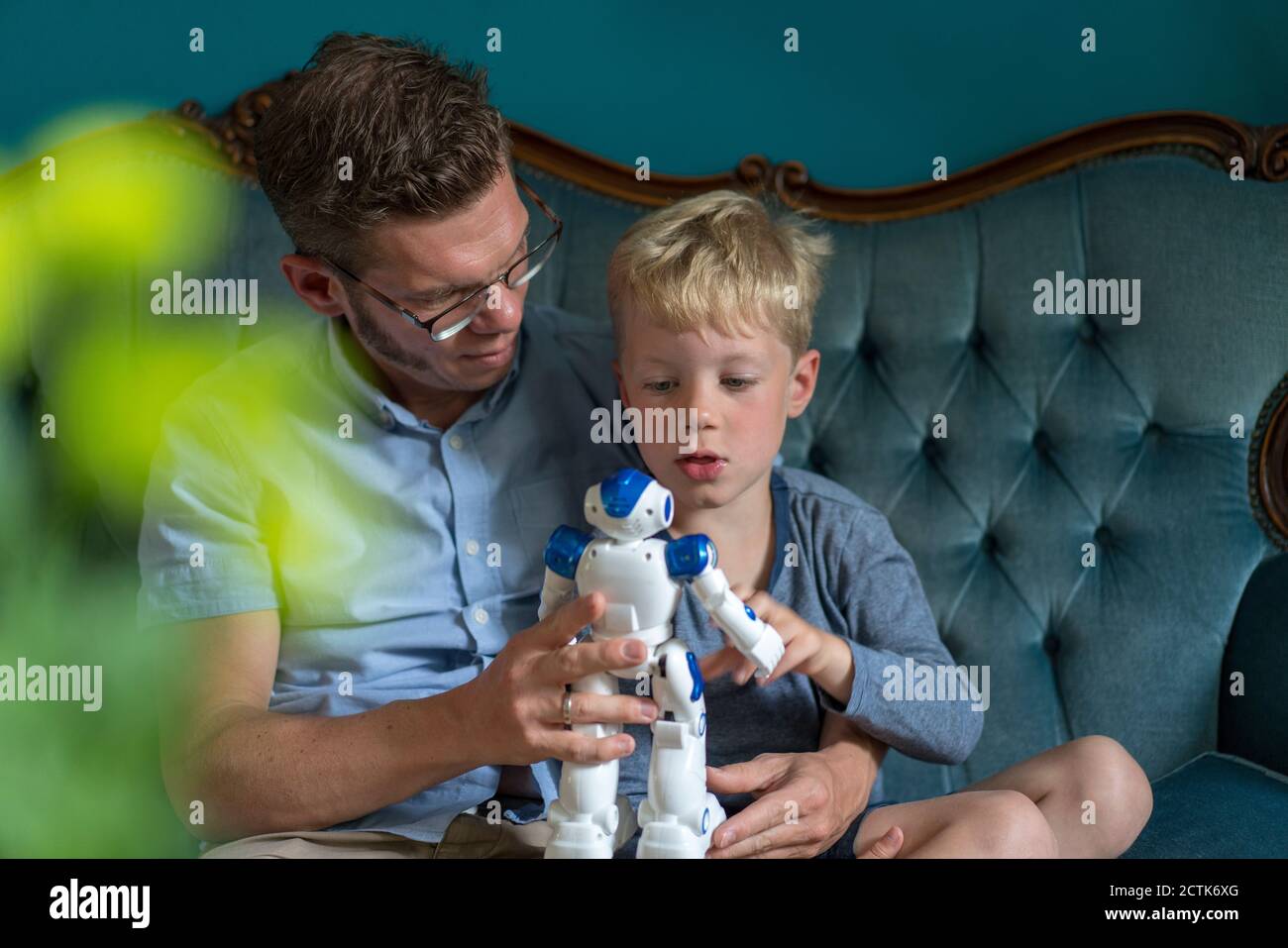 Cute blond boy looking at robot held by father while sitting on sofa in ...