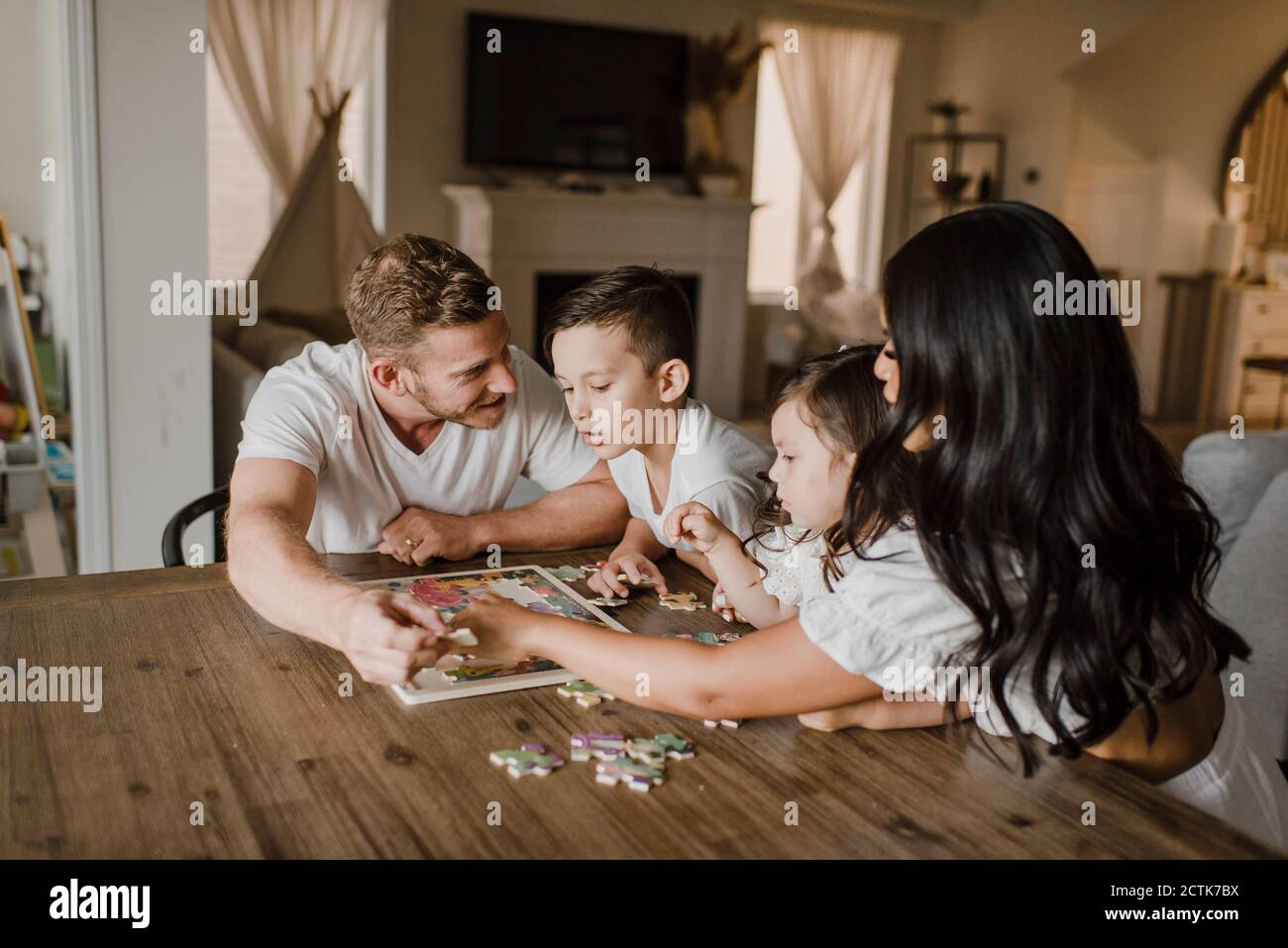 Smiling parents solving jigsaw puzzle with kids over table at home ...
