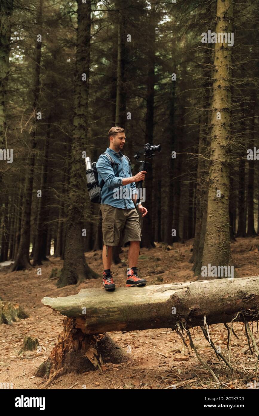 Man filming with camera and gimbal while standing on log against trees ...