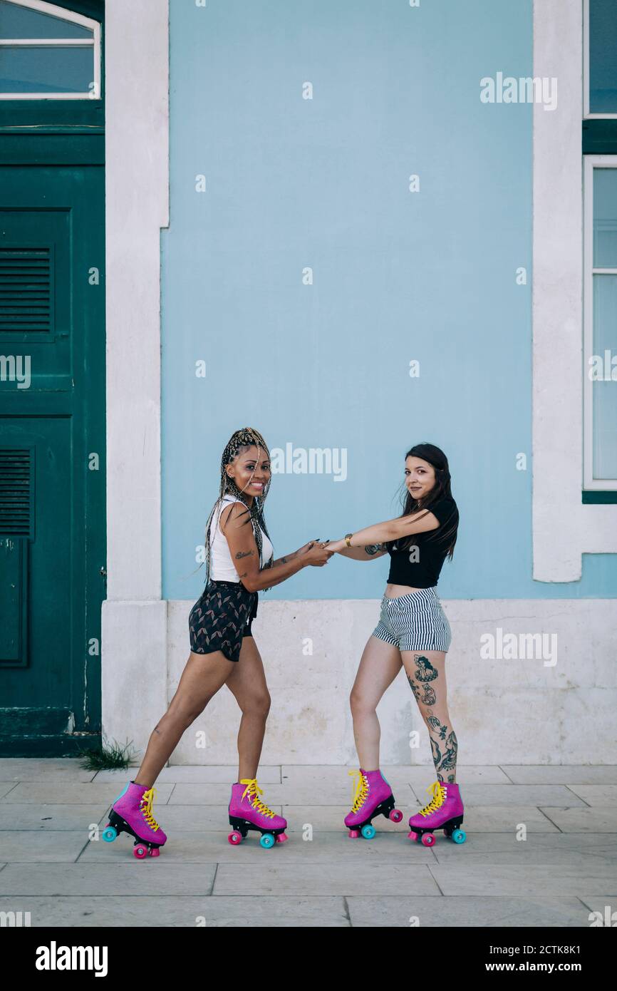 Friends holding hands while roller skating on footpath by wall in city ...