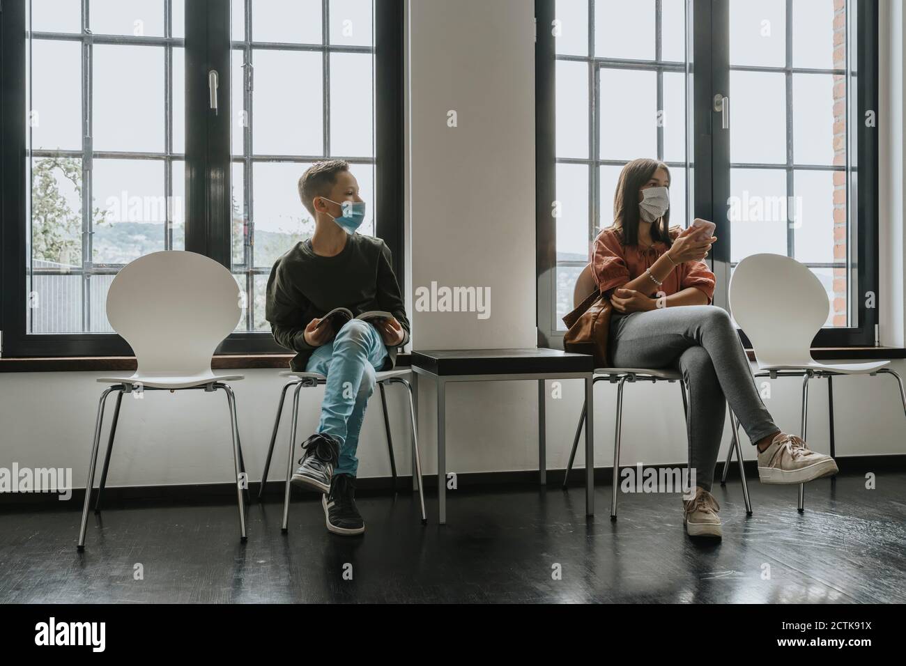 Teenage girl and boy wearing masks while sitting on chairs in waiting ...