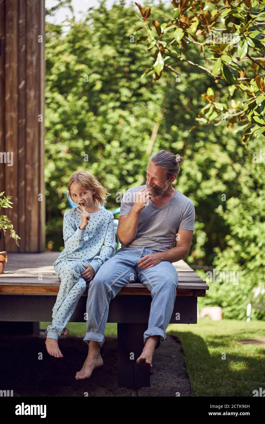 Father and daughter brushing teeth while sitting outside house in yard ...