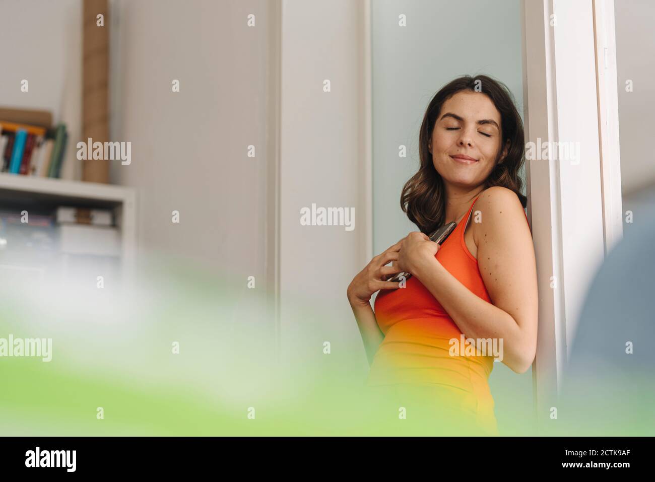 Beautiful woman hugging mobile phone while leaning on pillar at home ...