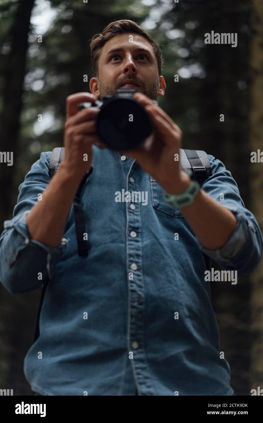 Serious male hiker holding camera looking away while standing in forest ...