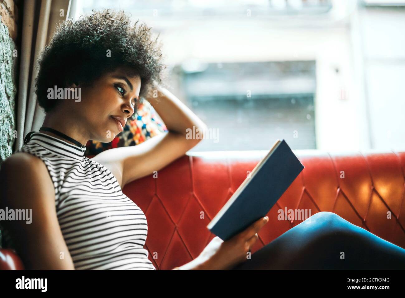 Mid adult woman with curly hair reading book while relaxing on sofa in ...