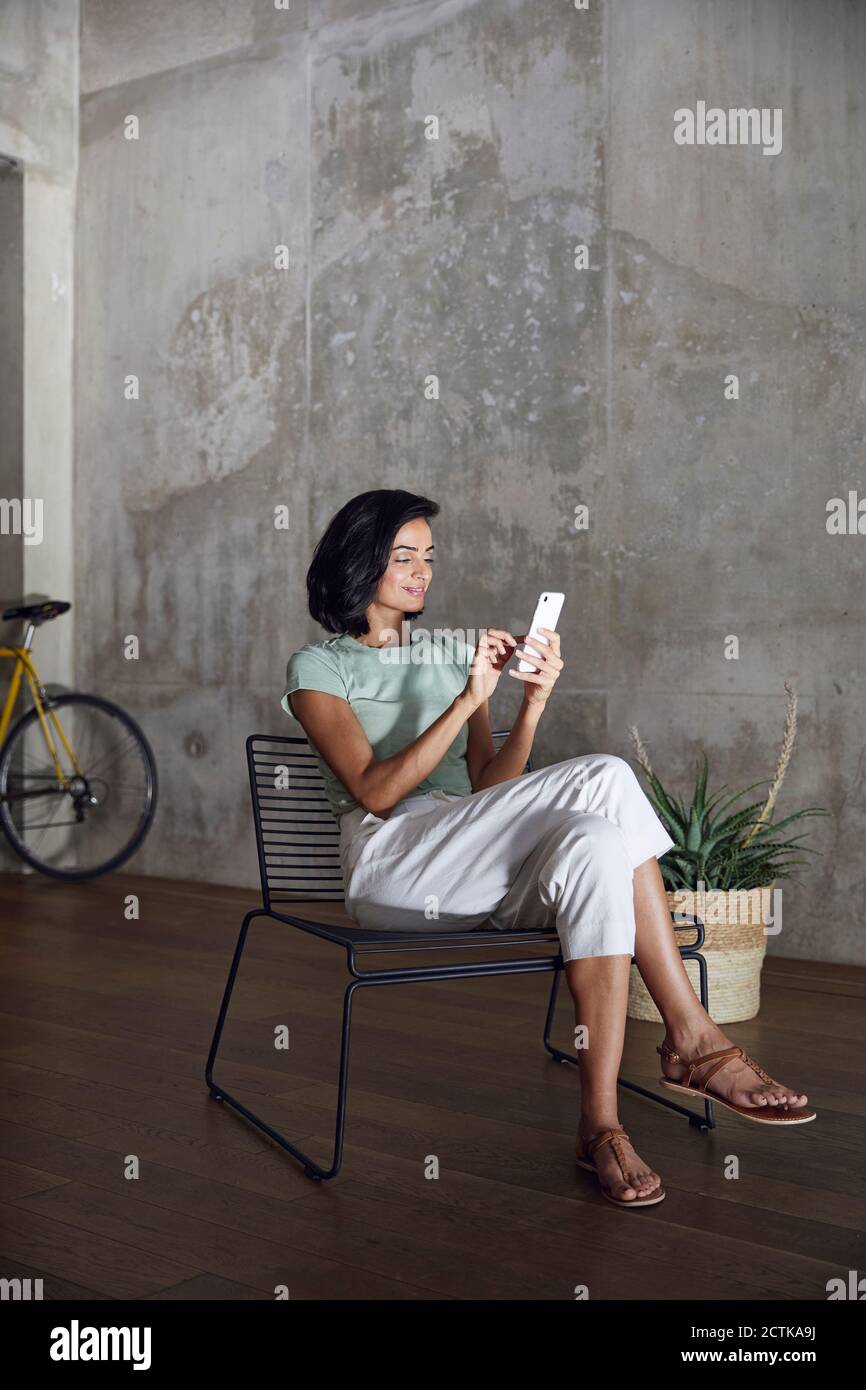 Businesswoman using mobile phone while sitting on chair against wall in ...
