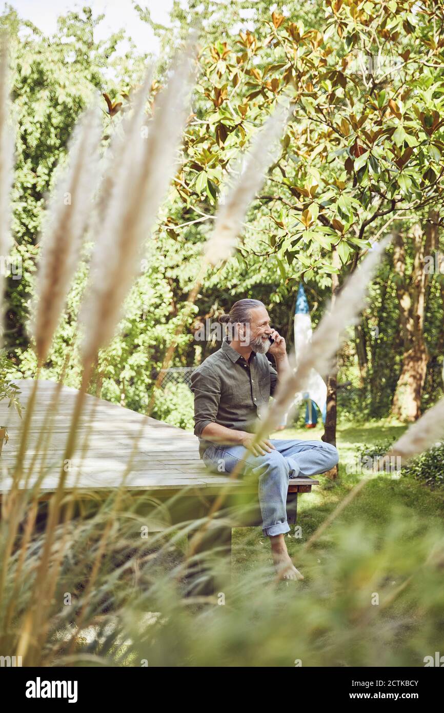 Mature man talking over smart phone while sitting against trees in yard ...