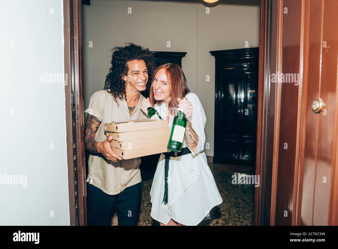 Cheerful friends with beer bottle and pizza boxes at entrance of home ...