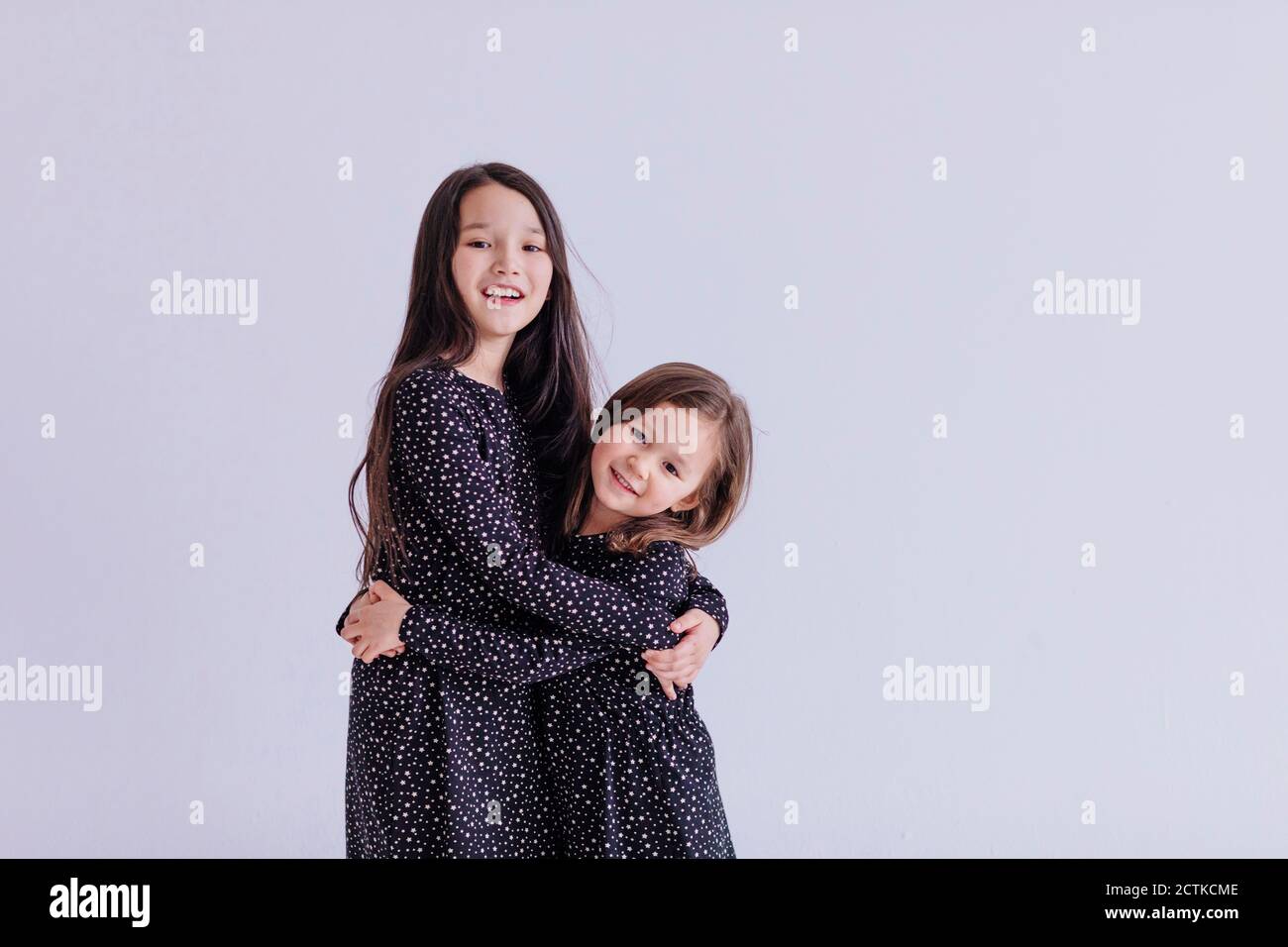 Smiling sisters embracing while standing against white background in ...