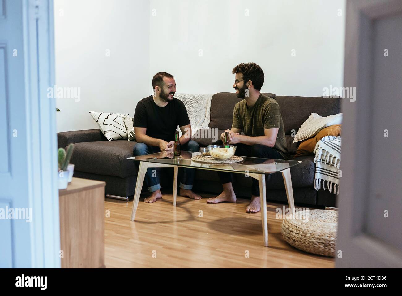 Male friends talking while sitting on sofa in living room seen through ...