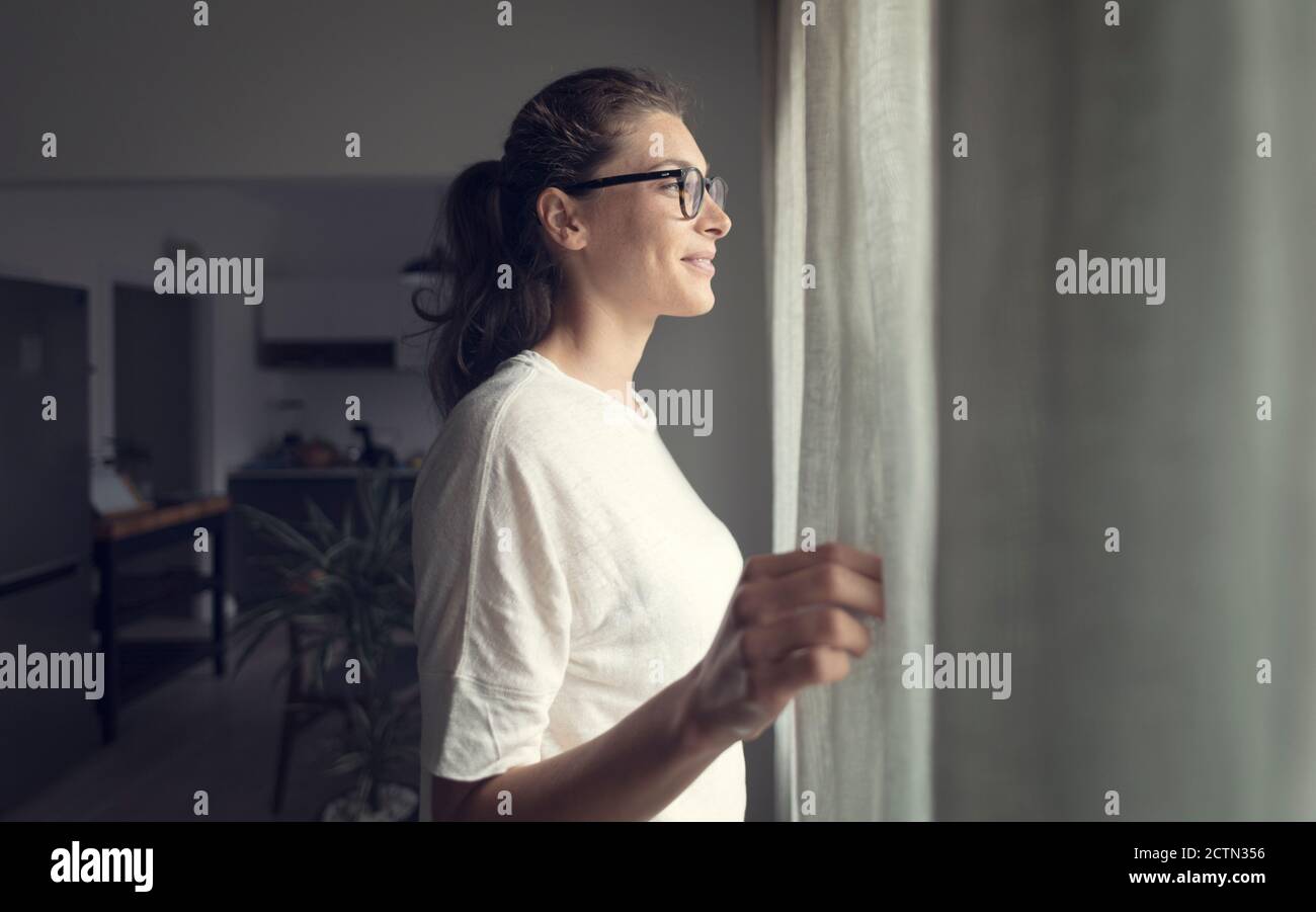 Happy woman with glasses standing next to a window and opening curtains ...