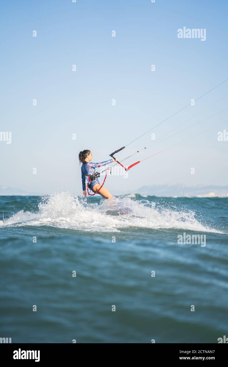 Side view of fit female kite surfer in swimsuit ridding waves in sea on ...