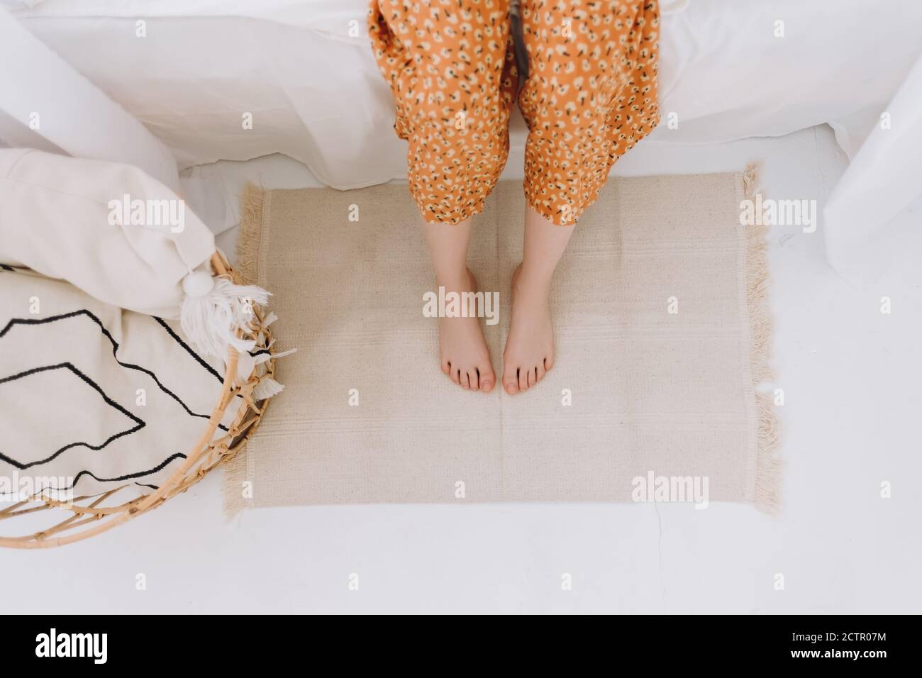 Young woman sitting on the bed put her feet on a rug made of natural ...