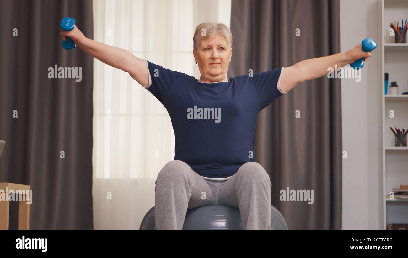 Old woman exercising on fitness ball in living room. Old woman lifting ...