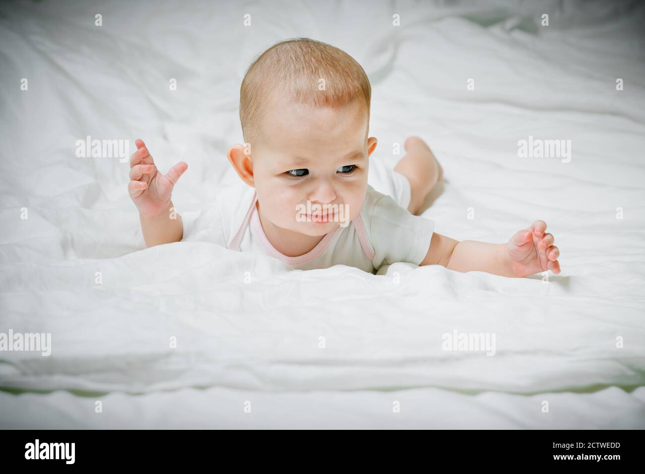 Sad and disappointed baby girl in white clothes is lying on a white bed ...