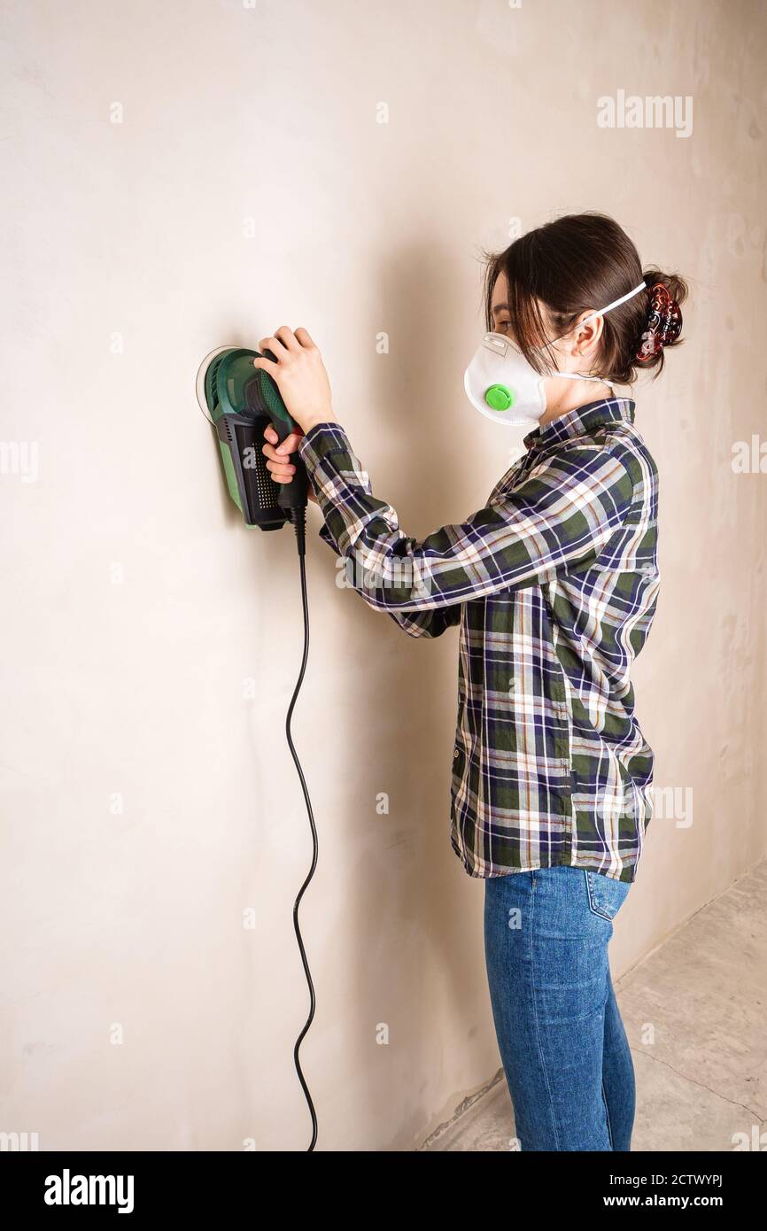 Woman in protective mask working with electric sander to smooth plaster ...