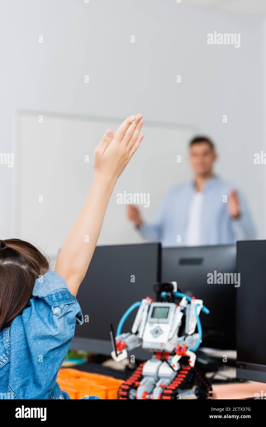 Selective focus of schoolgirl with raised hand sitting near robot and ...