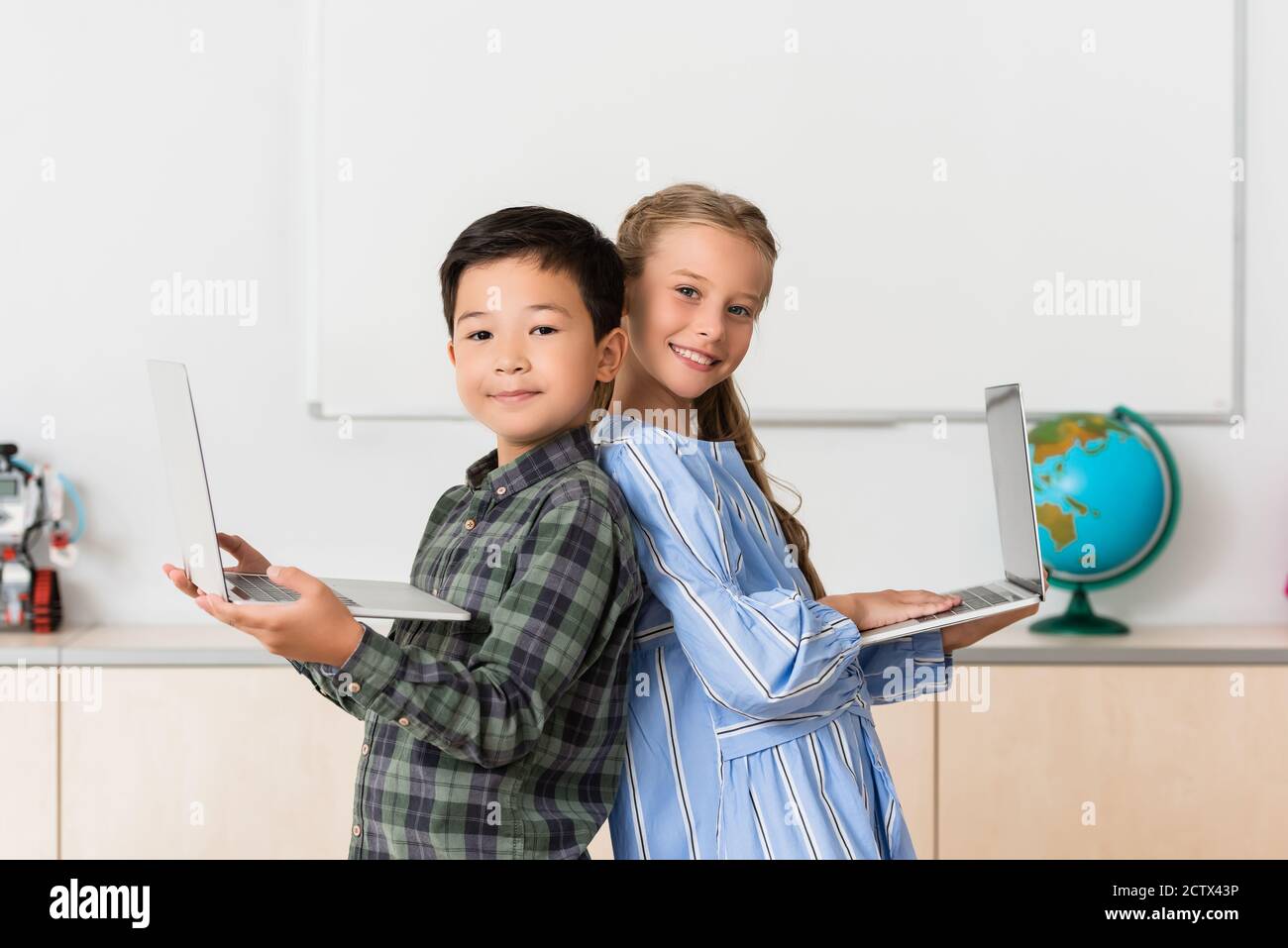 Multicultural pupils holding laptops and looking at camera in classroom ...