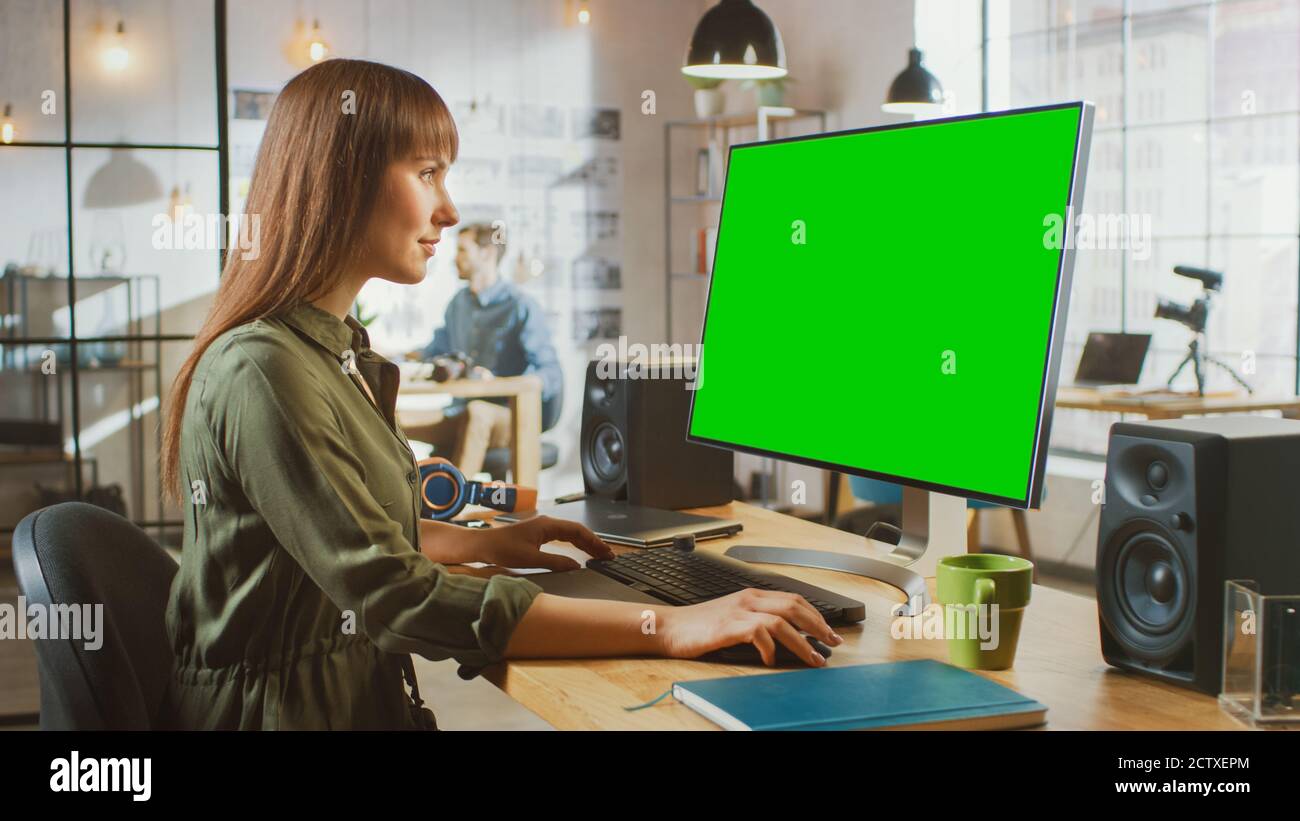 Beautiful Brunette Female Video Editor Works on Her Personal Computer with Big Green Screen Mock ...