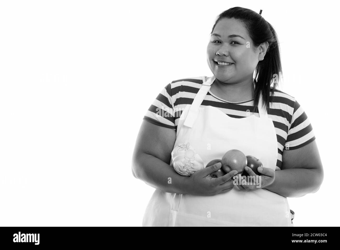 Studio shot of young happy fat Asian woman smiling while holding fruits ...