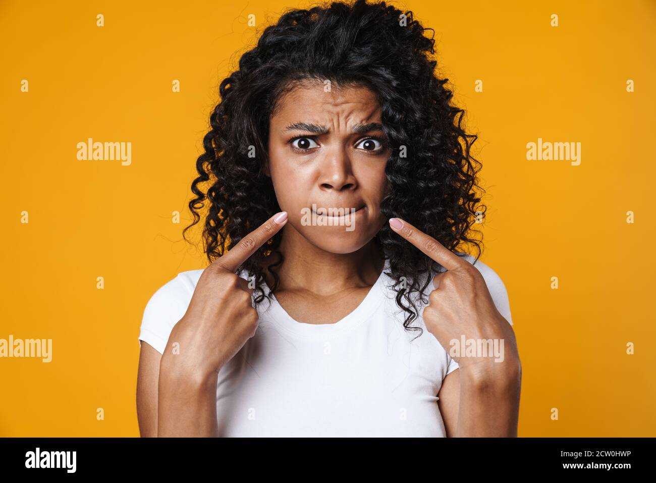 Image of confused african american woman pointing fingers at her cheeks ...
