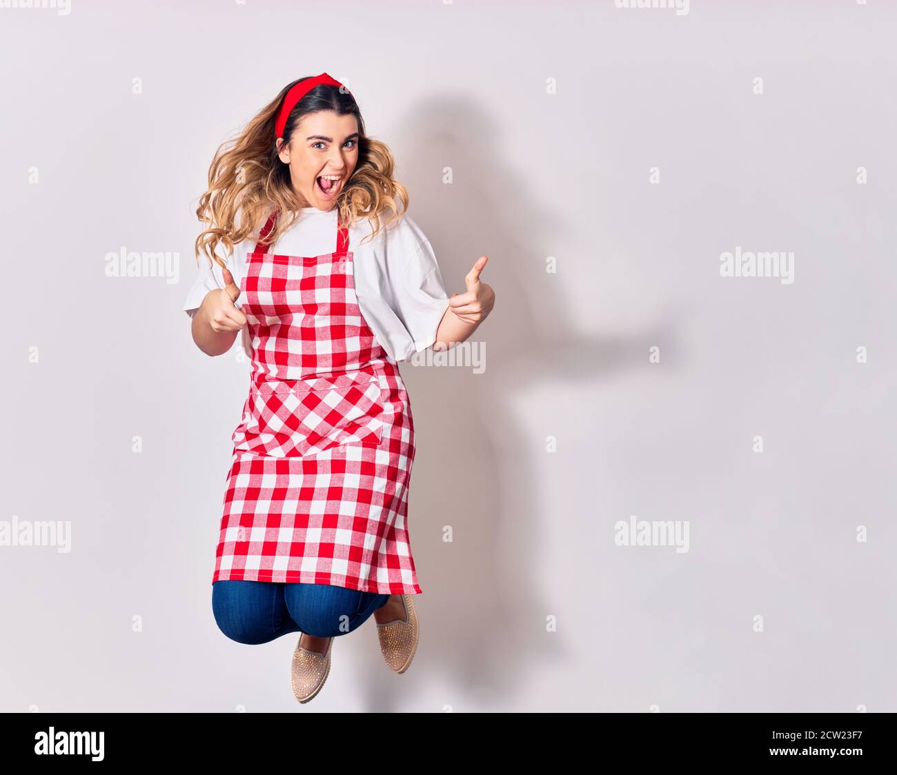 Young beautiful blonde cook woman wearing apron smiling happy. Jumping ...