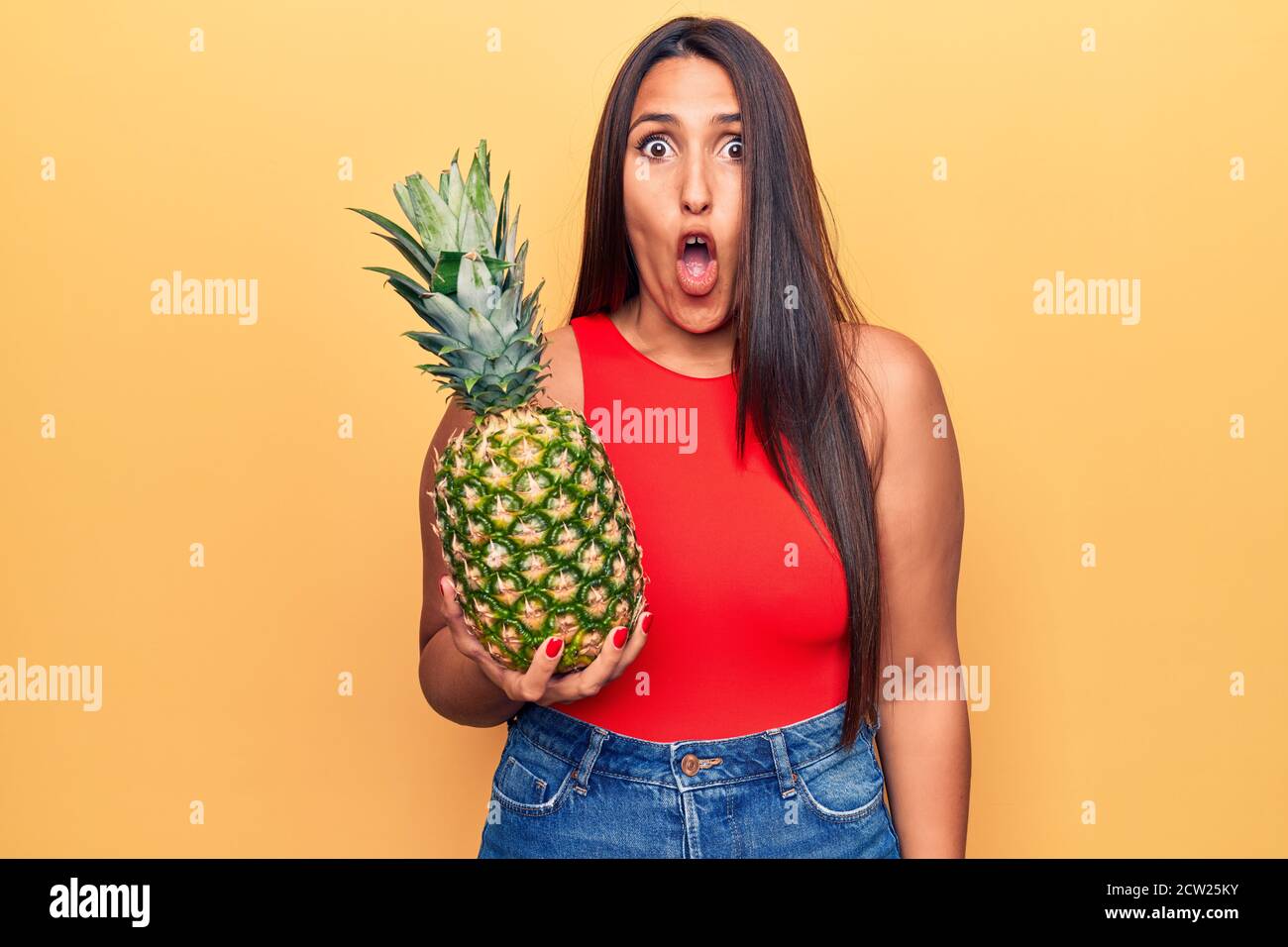 Young beautiful brunette woman holding pineapple scared and amazed with ...